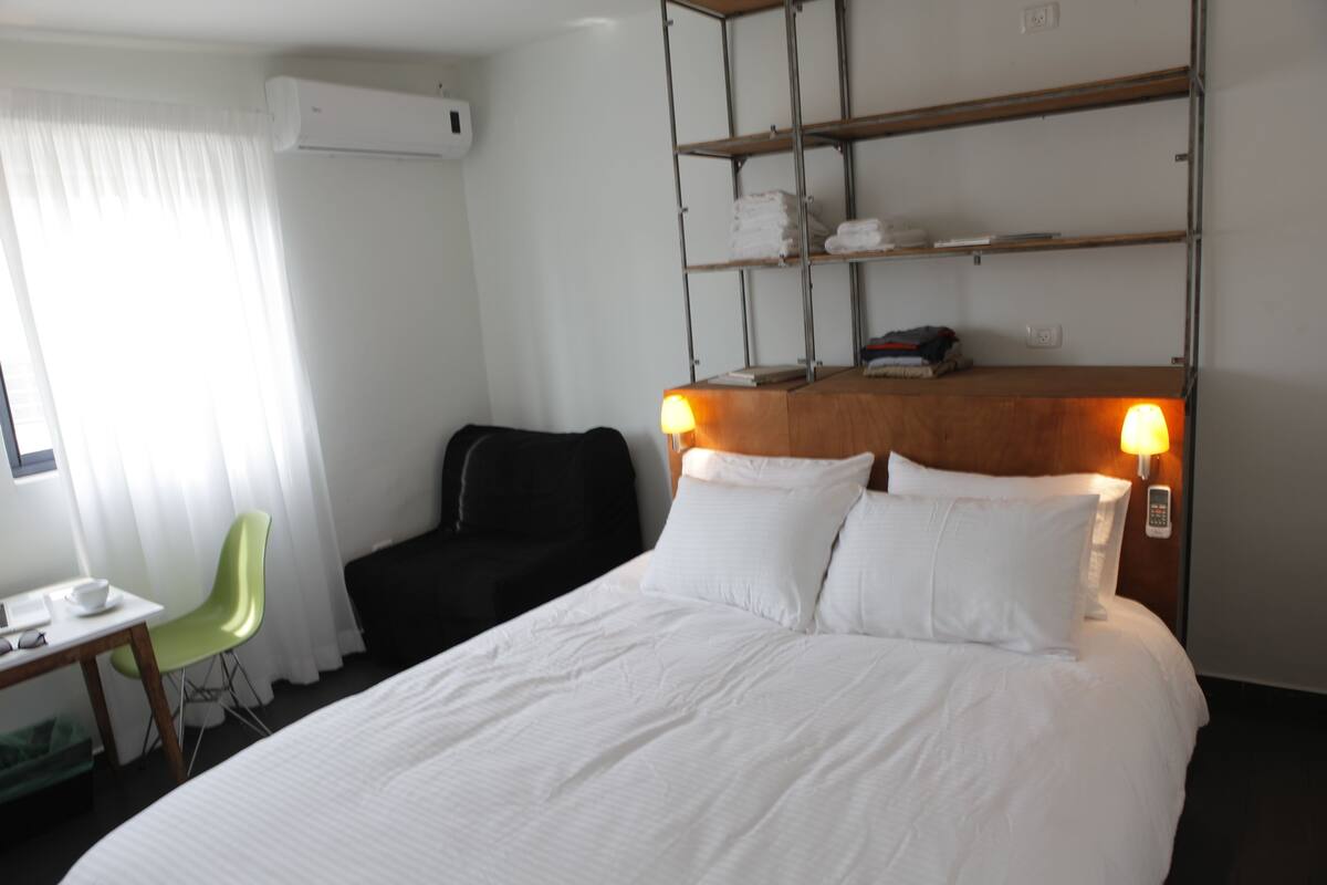 Quarto de hotel com uma cama, uma mesa, uma cadeira, uma prateleira com toalhas e um ar condicionado fixado na parede.