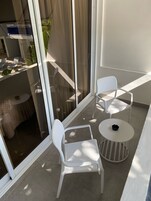 Uma varanda com duas cadeiras brancas e uma mesa redonda pequena, uma porta de vidro e vista para um quarto com um tapete estampado.