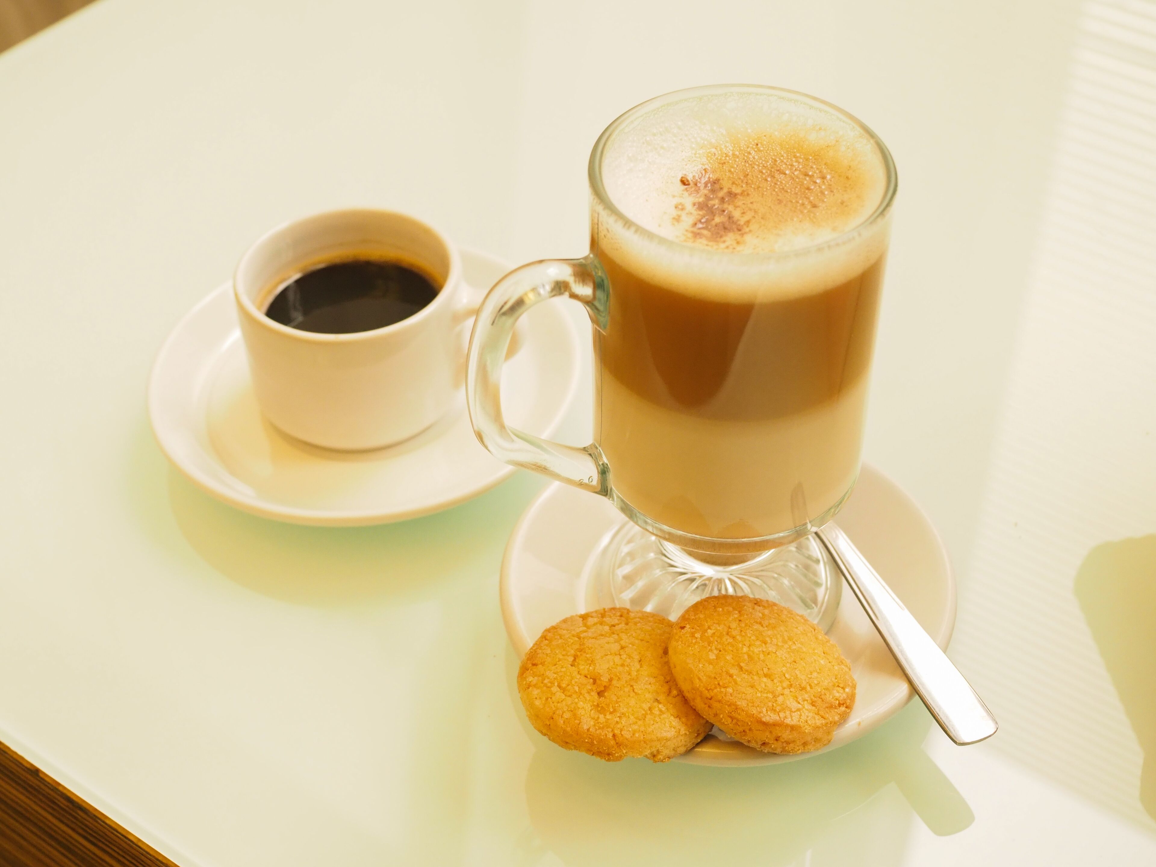 Uma chávena de vidro transparente com café, uma pequena chávena de café preto e duas bolachas sobre um pires branco.