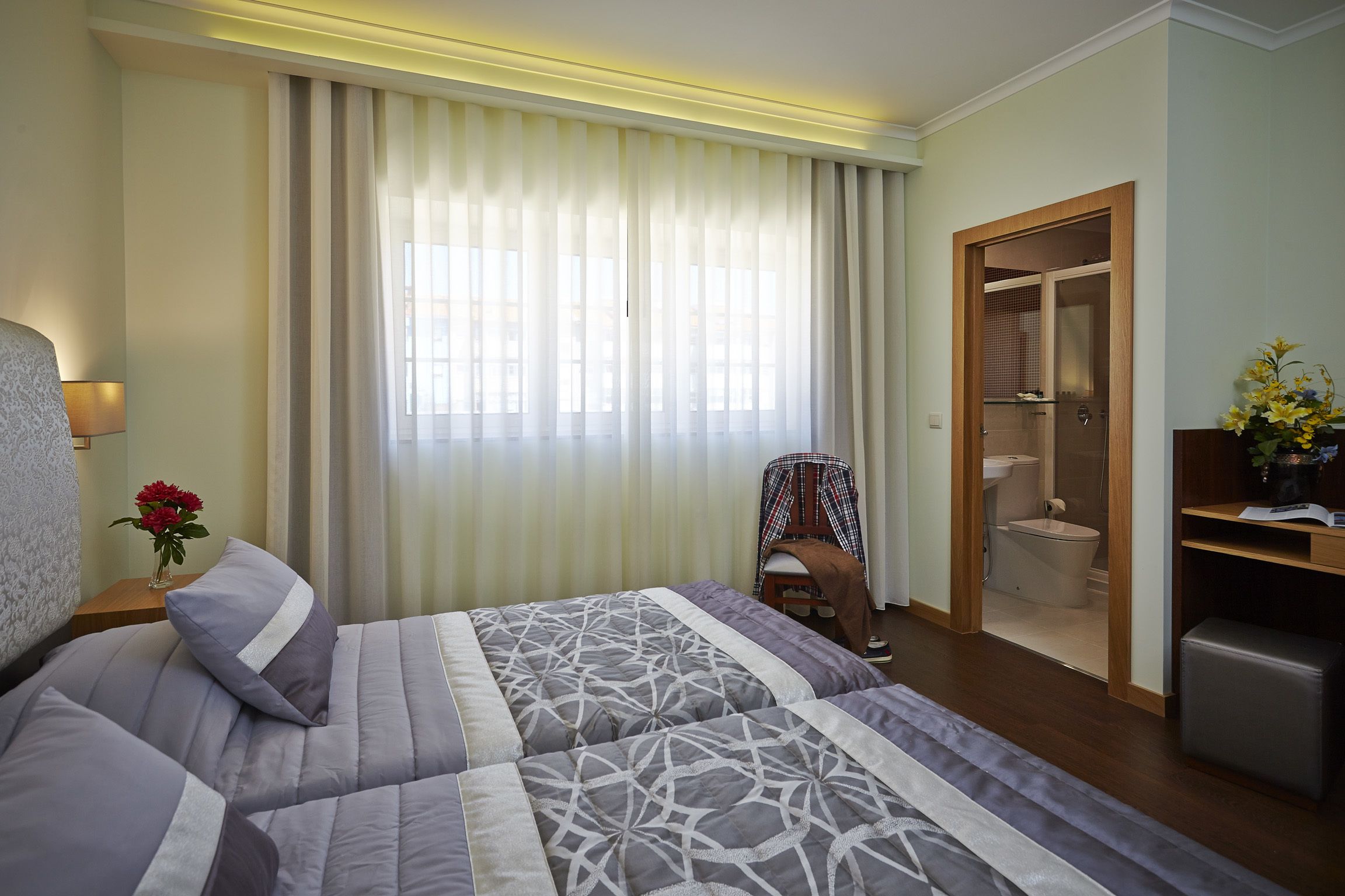 Quarto de hotel com cama grande, escrivaninha de madeira, cadeira, guarda-roupa e banheiro visível através de uma porta aberta.