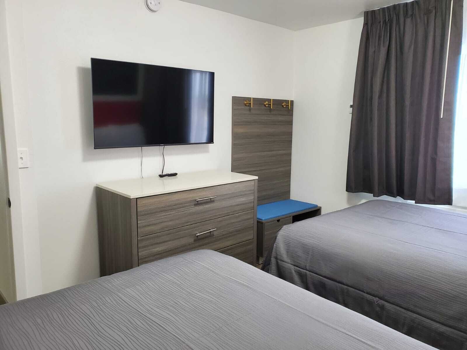 Quarto de hotel com televisão de tela plana na parede, cómoda de madeira com gavetas, duas camas com roupa de cama cinzenta e uma janela com cortinas escuras.
