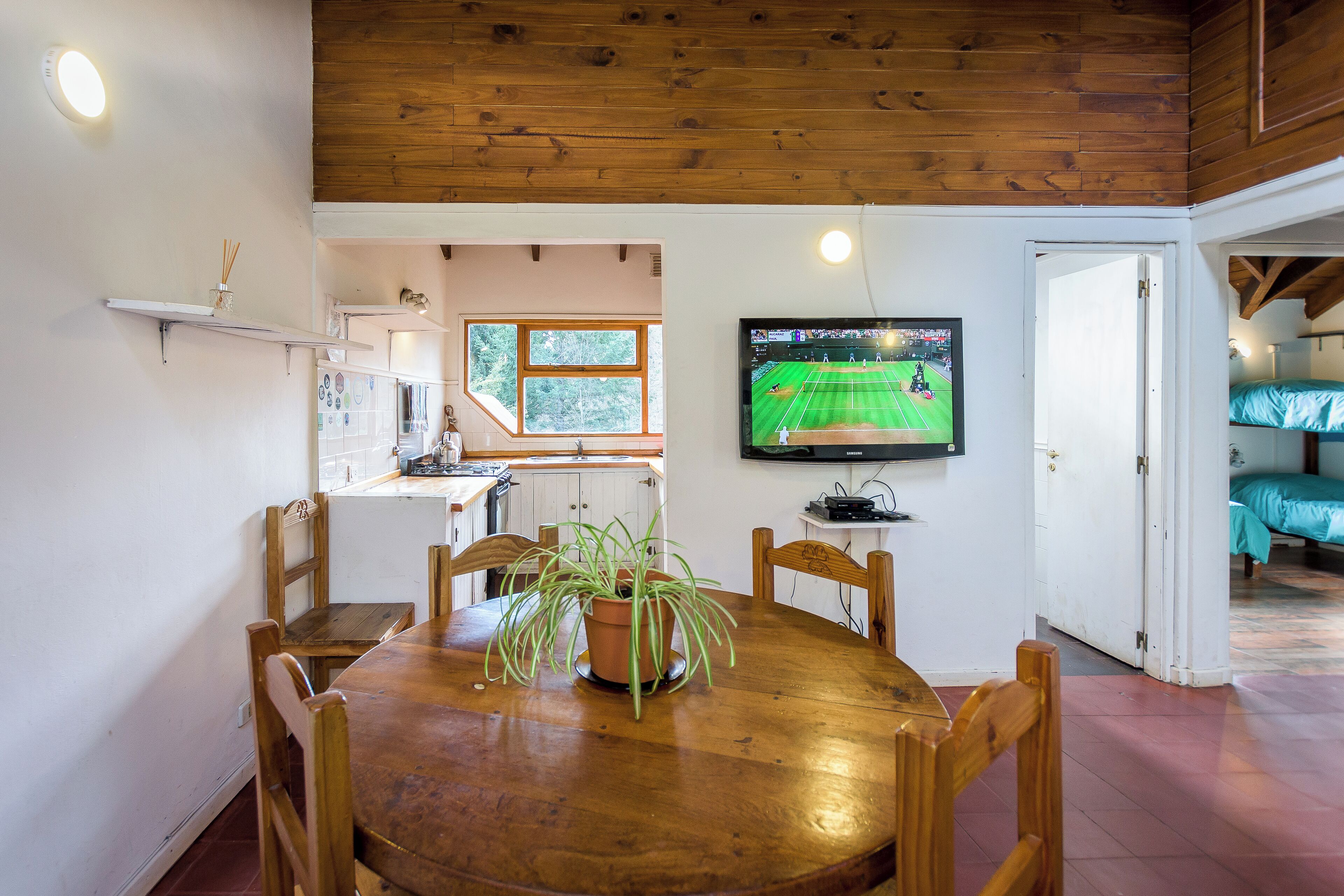 Uma área de refeições com mesa e cadeiras de madeira, um vaso com planta, uma televisão exibindo uma partida de tênis e uma cozinha ao fundo.