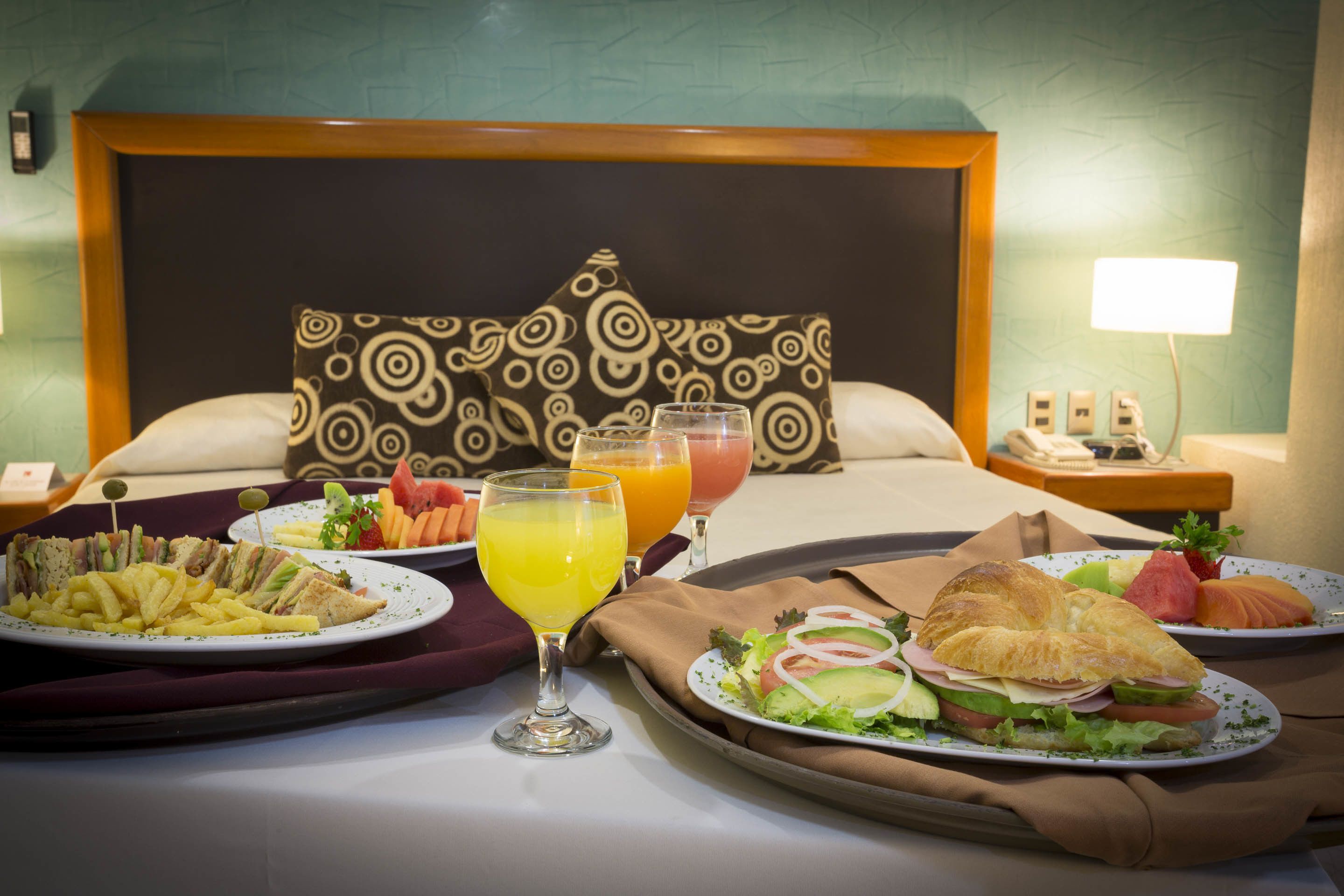 Disposição de café da manhã com sanduíches, frutas e sucos sobre uma cama de hotel.