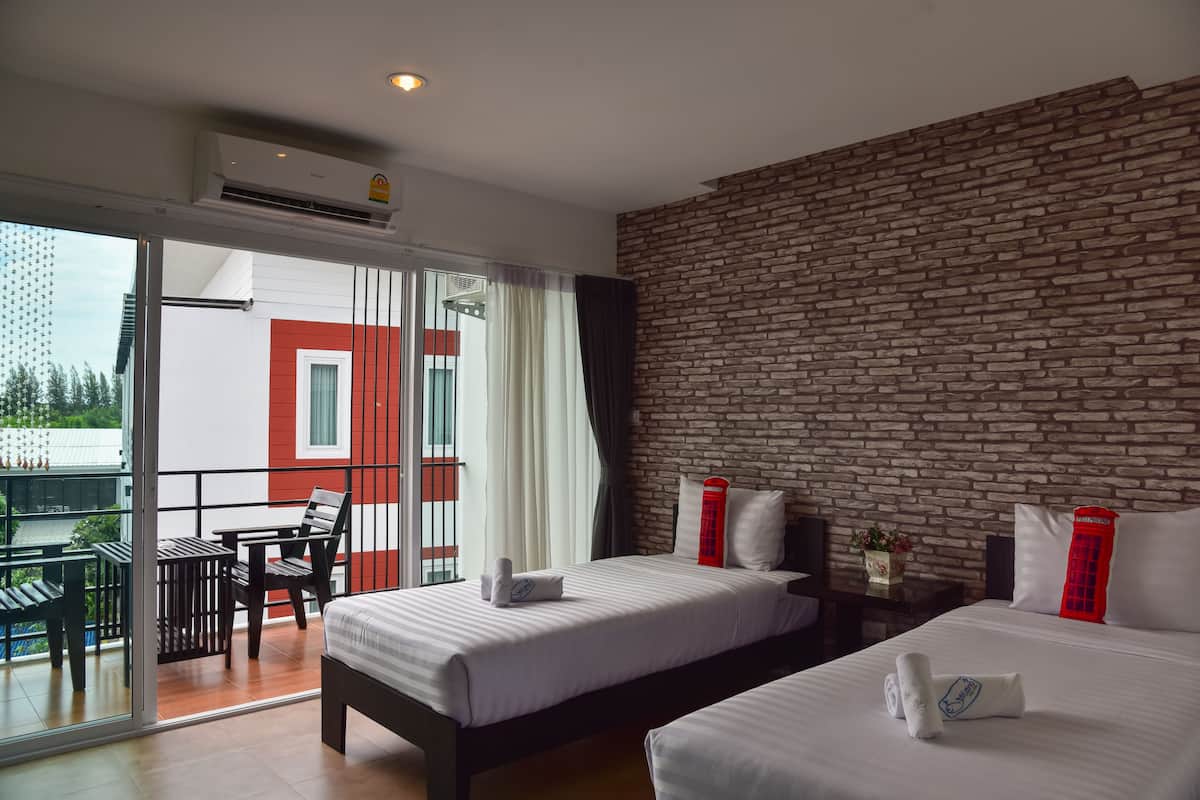 Quarto de hotel com duas camas, parede de tijolos, varanda com mesa e cadeiras e vista para um edifício com fachada vermelha e branca.