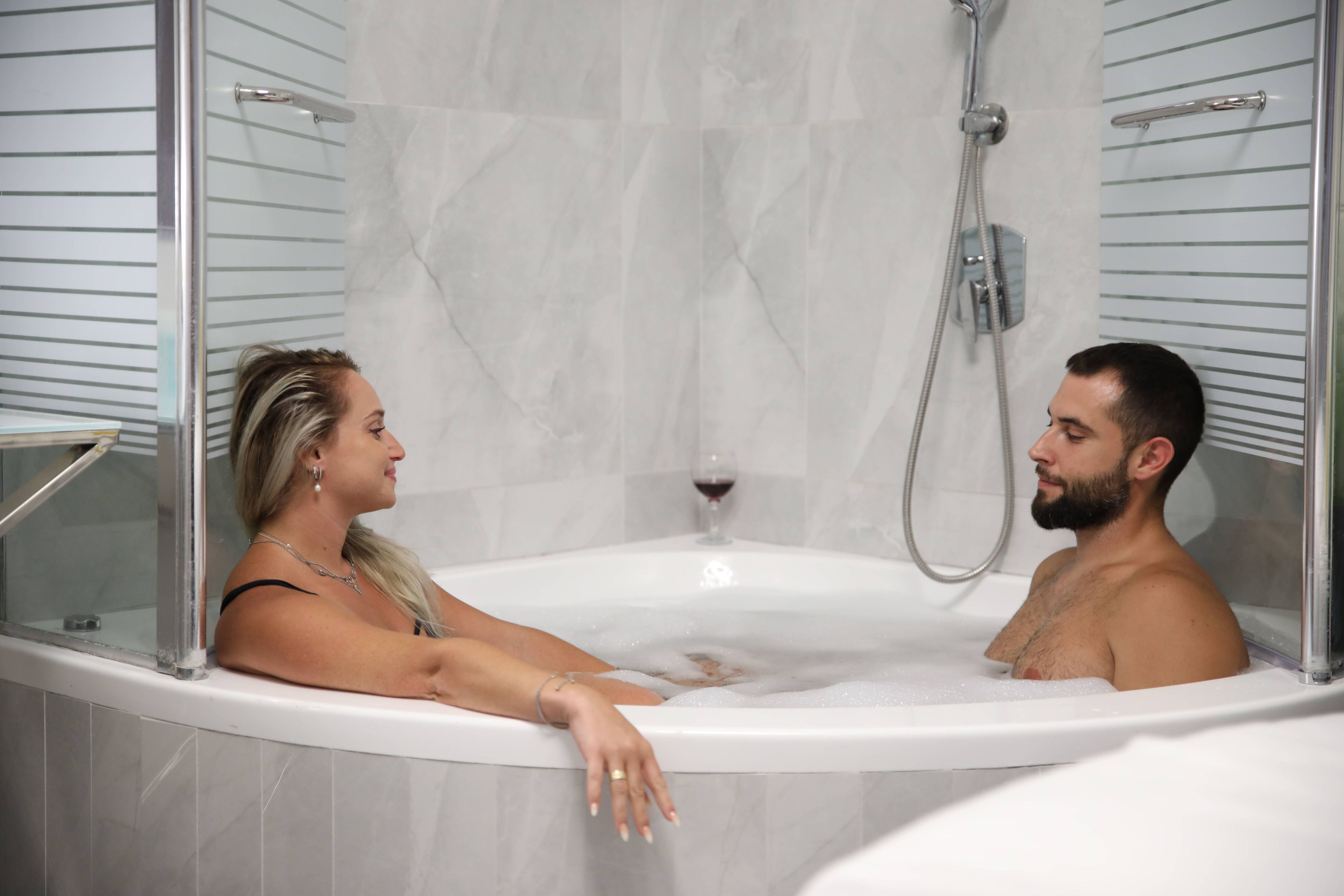 Um casal em um banheiro moderno com banheira de vidro, chuveiro manual e uma taça de vinho na borda.