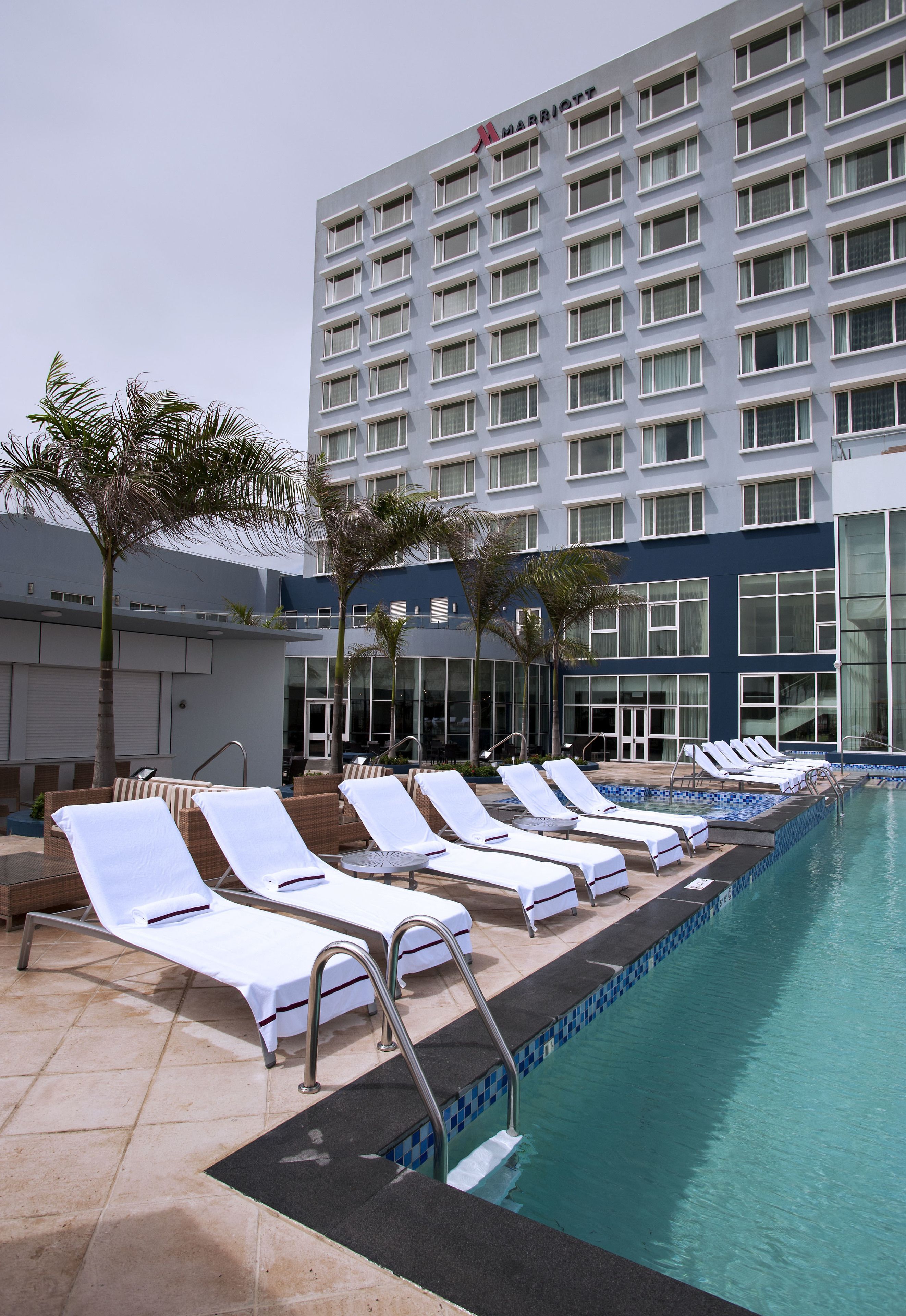 Área de piscina de hotel com espreguiçadeiras, palmeiras e um edifício grande ao fundo.