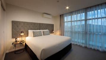 Quarto de hotel com uma cama grande, uma mesinha de cabeceira com luminária, telefone e ar-condicionado fixado na parede.