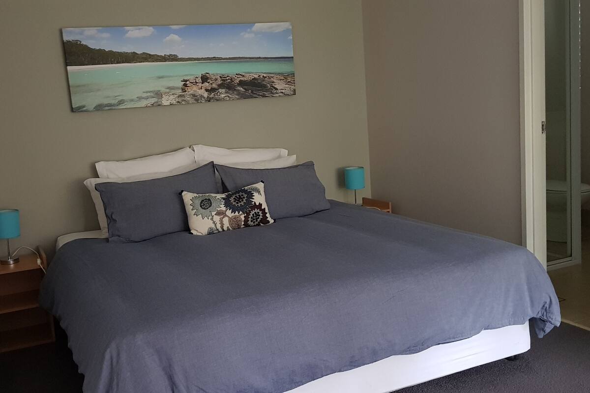 Quarto com cama, mesa de cabeceira, luminária, pintura de uma praia e porta do banheiro.