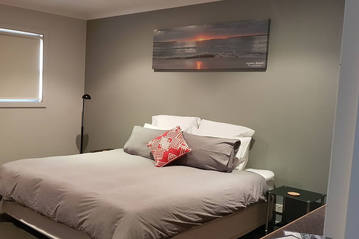 Quarto com cama, cadeira, mesa de cabeceira, luminária, janela e um quadro na parede.