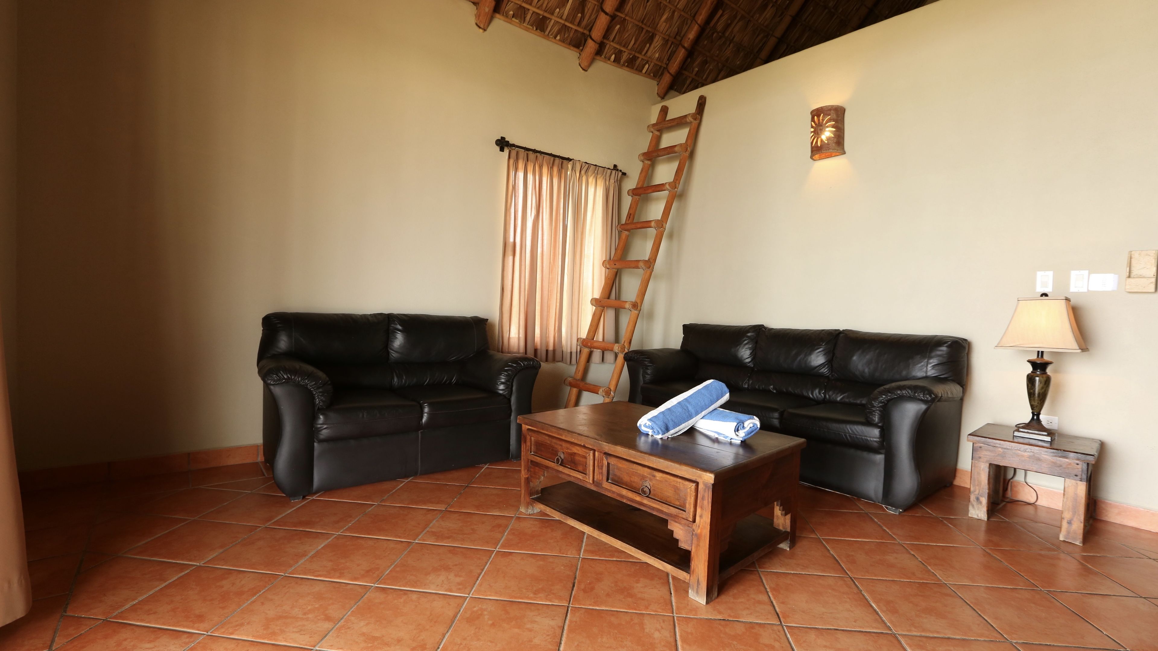 Uma sala de estar com dois sofás de couro preto, uma mesa de centro de madeira, uma escada e um abajur sobre uma mesinha lateral.