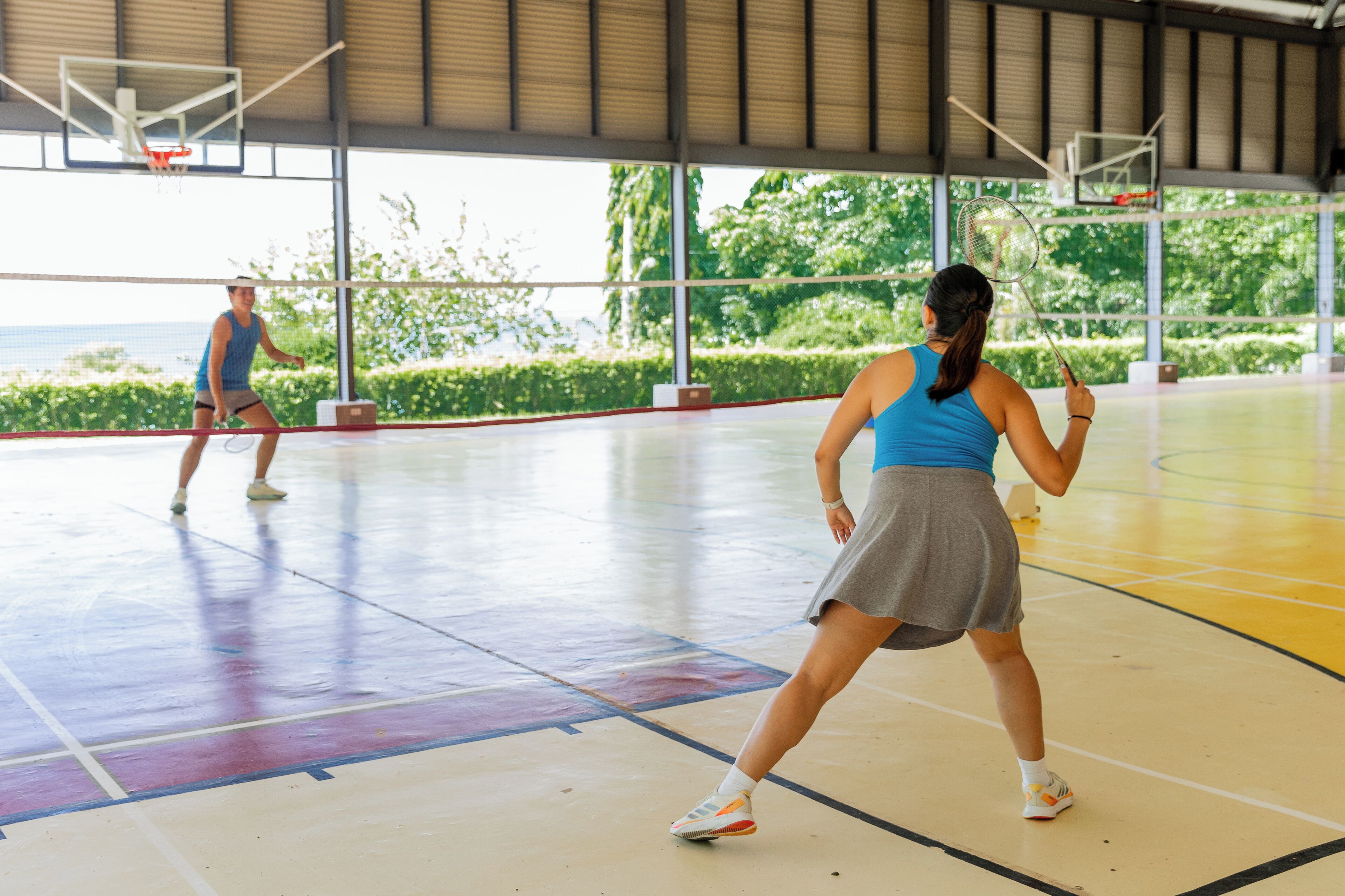 Uma quadra de basquete interna com dois jogadores, um de camiseta azul e saia cinza, e outro de camiseta azul e shorts.