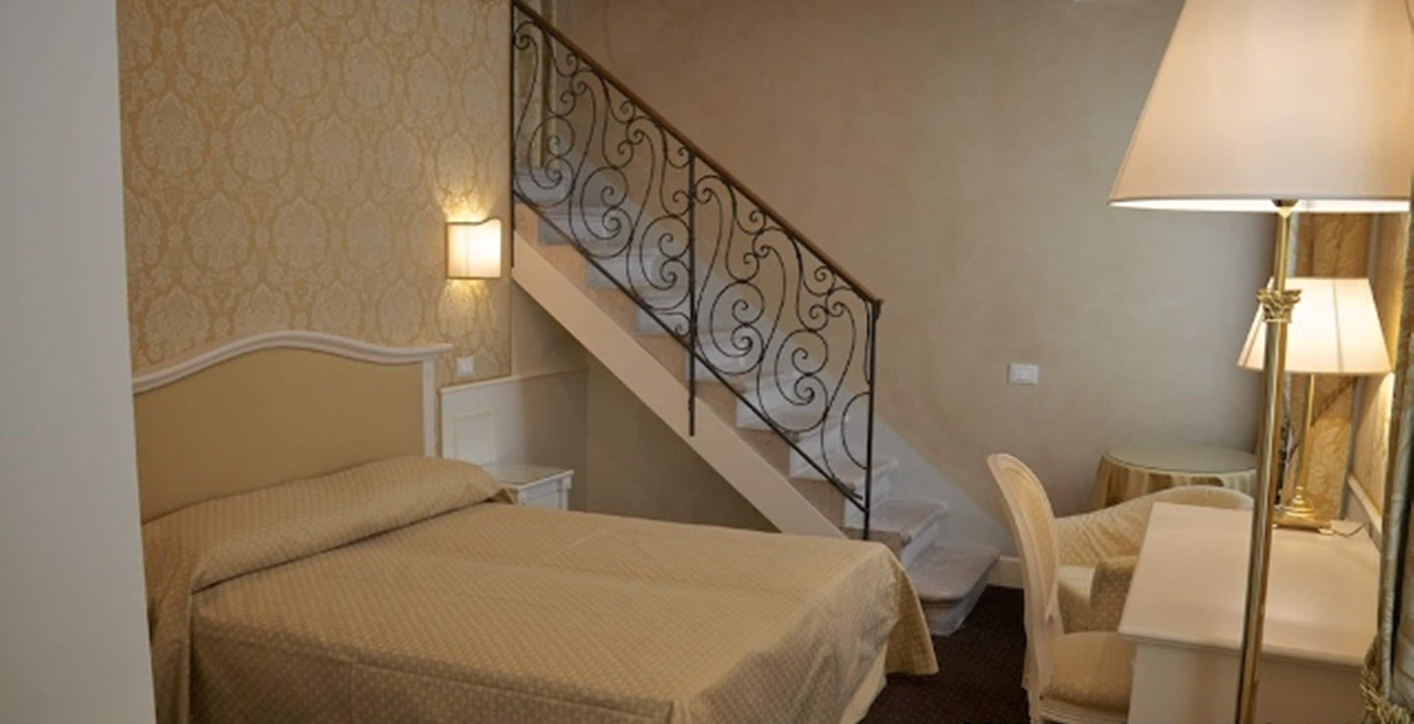 Quarto de hotel com cama, poltrona, escrivaninha, luminária e escada com corrimão decorativo.