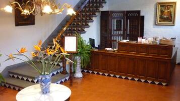 Uma área de recepção com um balcão de madeira, um vaso com flores, uma escadaria e um candelabro.