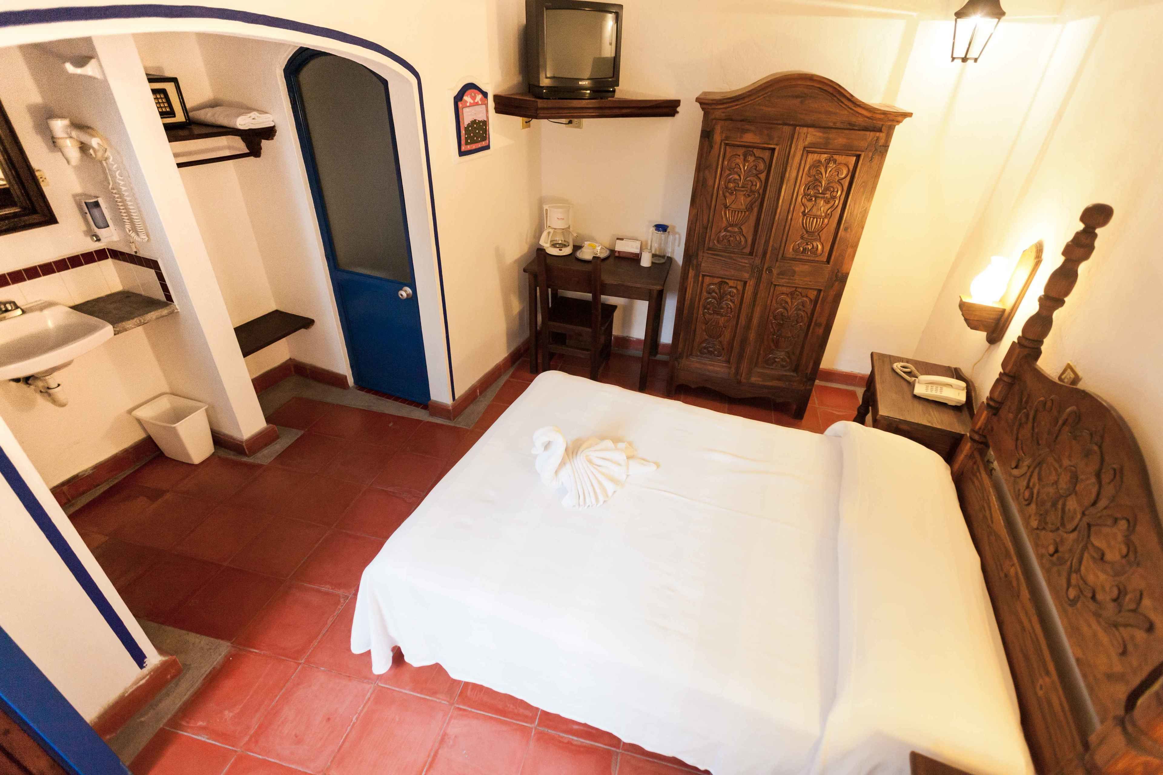 Quarto de hotel com cama, secretária, guarda-roupa, televisão, telefone e banheiro.