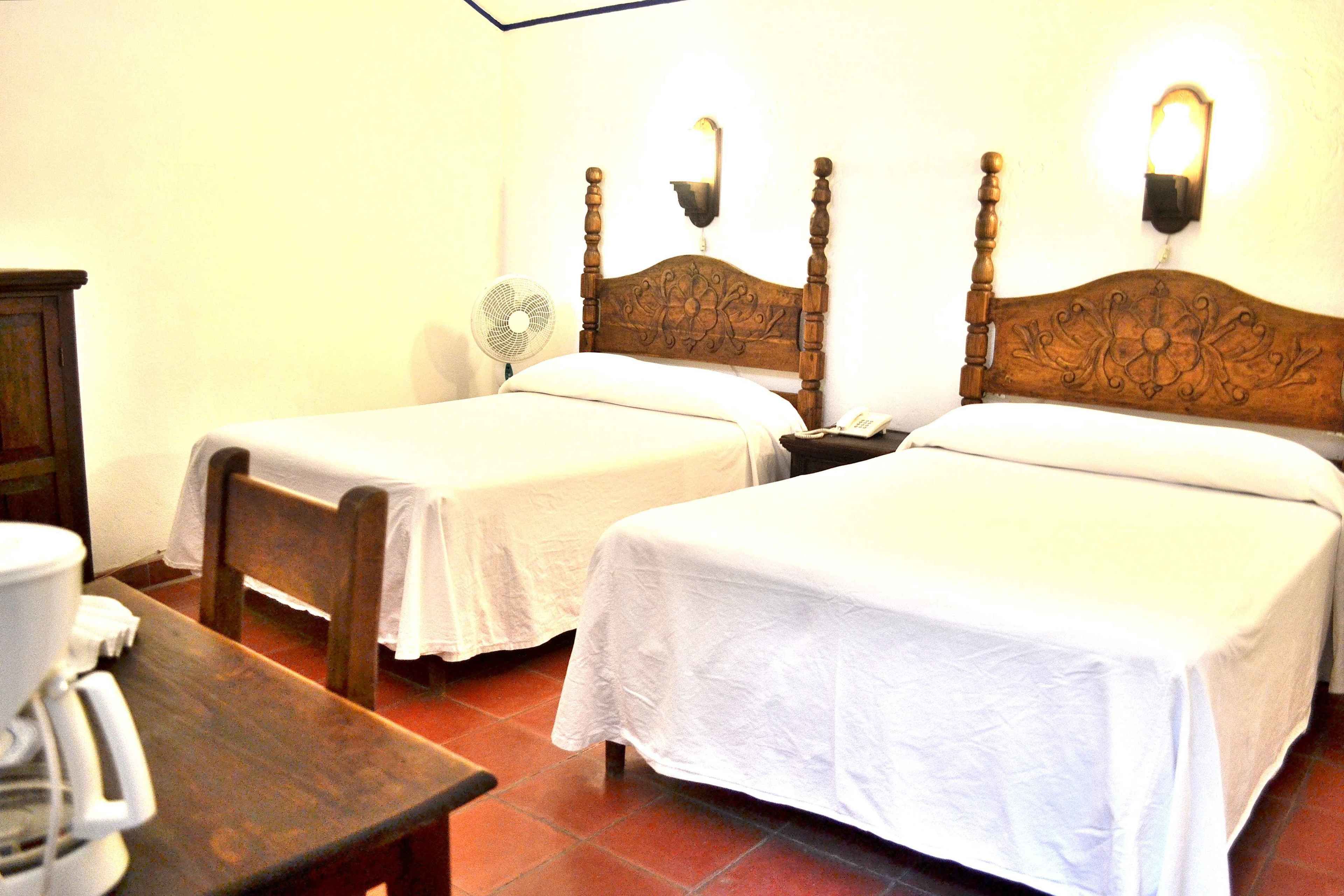 Quarto de hotel com duas camas de solteiro, uma mesa de madeira, uma cadeira, mesa de cabeceira com abajur e ventoinha fixada na parede.