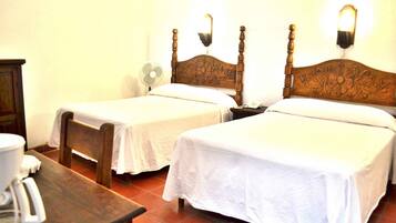 Quarto de hotel com duas camas de solteiro, uma mesa de madeira, uma cadeira, mesa de cabeceira com abajur e ventoinha fixada na parede.