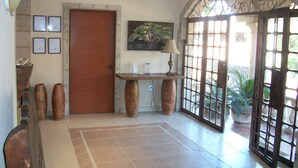 Um hall amplo com porta de madeira, piso de azulejos, uma mesa com abajur e uma janela grande com moldura metálica decorativa.