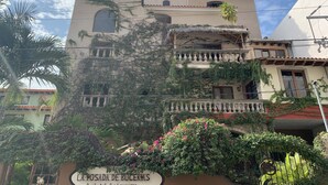 Um edifício com uma placa que diz "La Posada de Bucerias". Apresenta várias varandas com guarda-corpos, um telhado de palha no andar superior e é cercado por vegetação exuberante e plantas floridas.