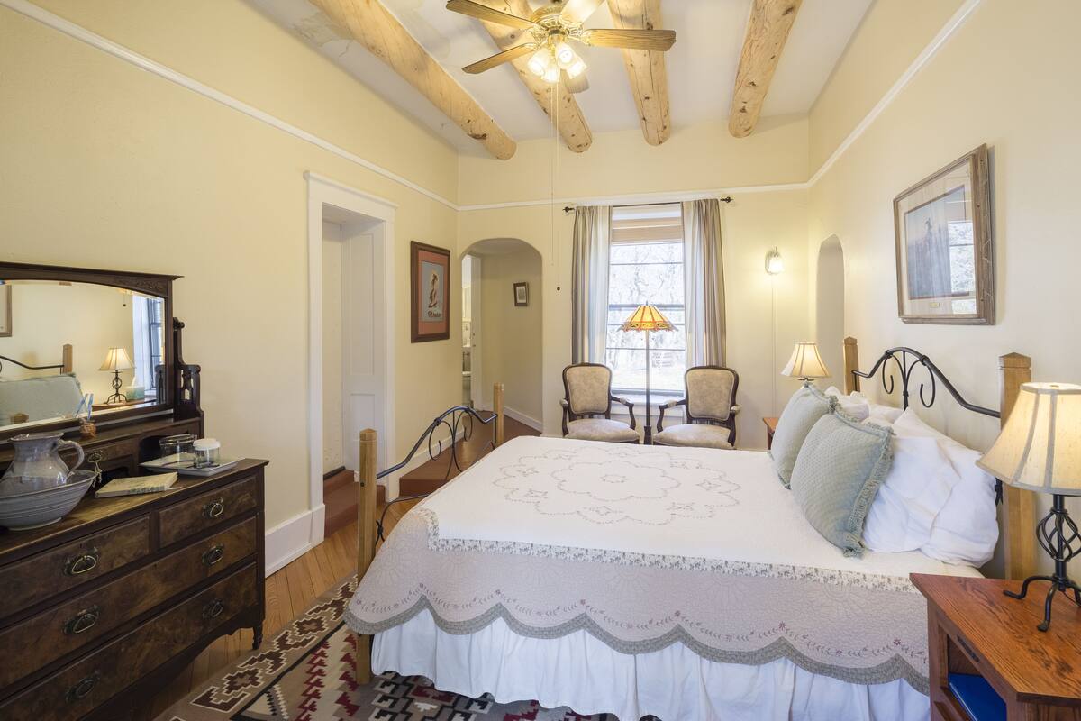 Quarto com cama grande, vigas de madeira no teto, cômoda, espelho e janela com cortinas.