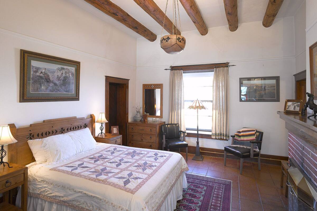 Quarto com uma cama grande, vigas de madeira, cômoda, cadeira, lareira e janela com cortinas.