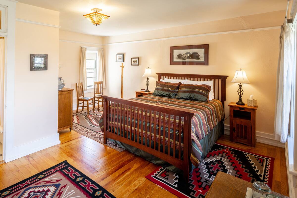 Quarto com cama de madeira, duas mesas de cabeceira com luminárias, uma cadeira, um tapete e uma porta que dá para o banheiro.
