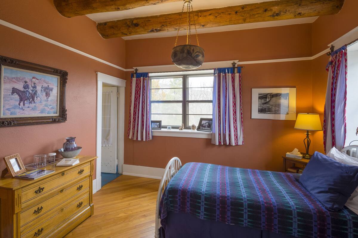 Quarto com cómoda de madeira, cama com colcha estampada, janela com cortinas e um quadro na parede.