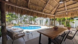 Uma área de piscina com telhado de palha, cadeiras de espreguiçadeira e uma mesa de madeira.