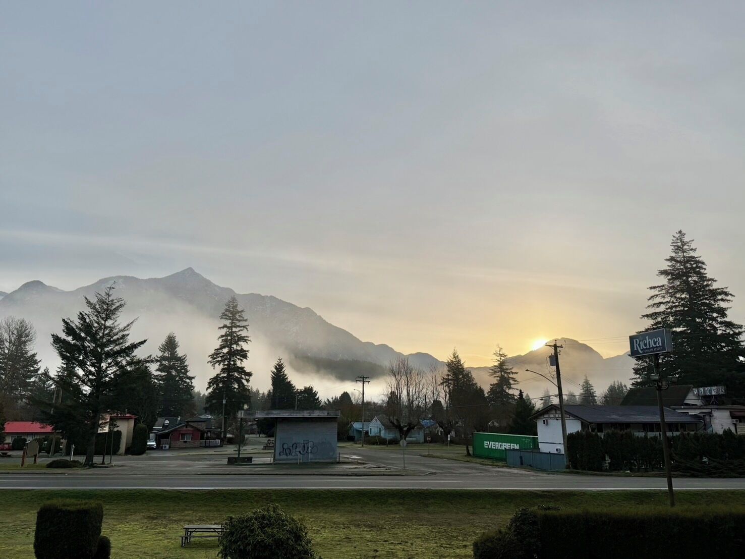 Vista de uma cordilheira com nascer ou pôr do sol, um letreiro para 'Ricca Restaurant' e um edifício com a inscrição 'Evergreen'.