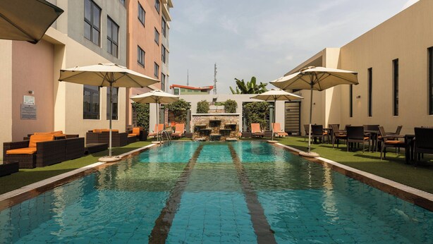 Piscina com espreguiçadeiras e guarda-sóis, rodeada por edifícios.