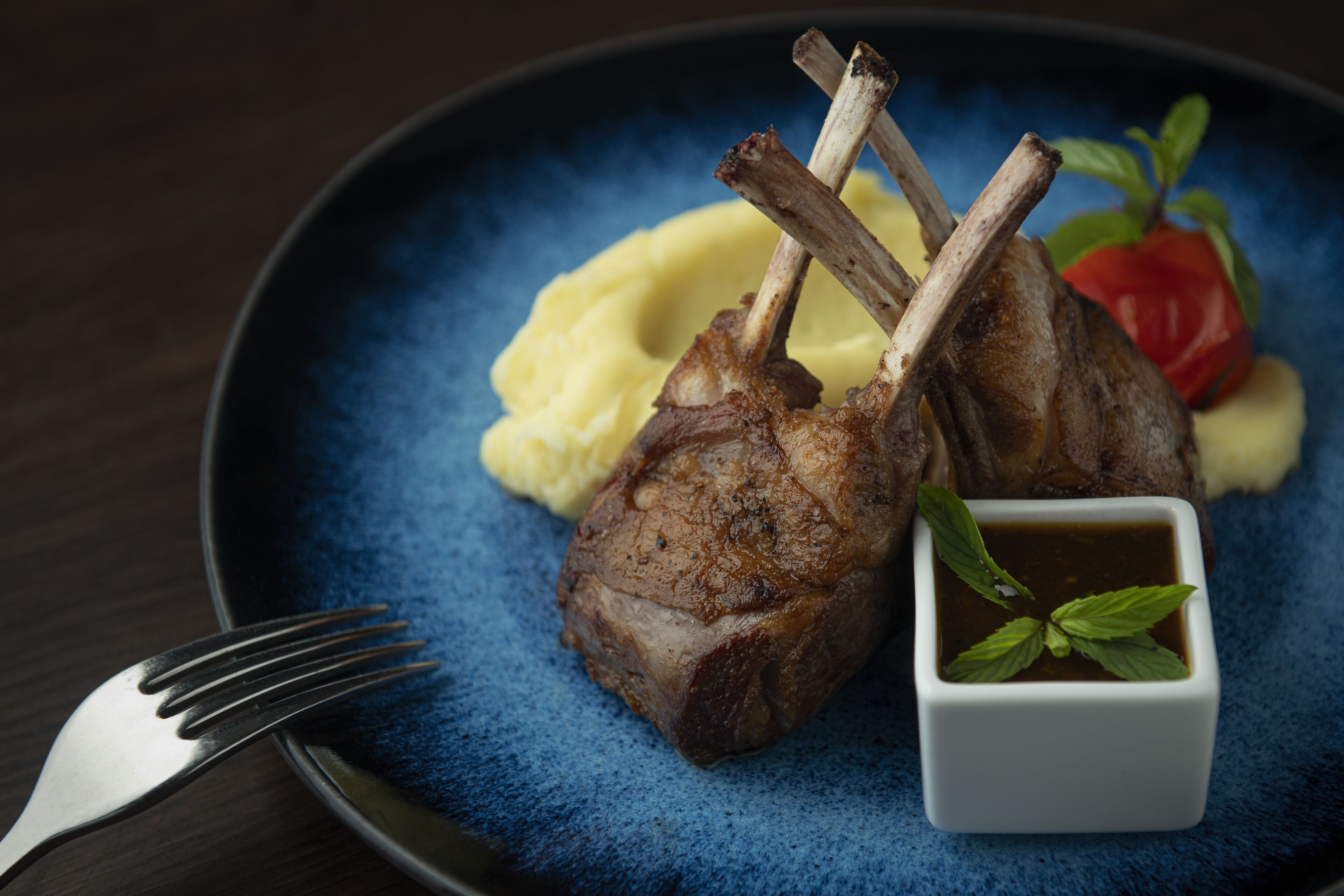 Prato com um bife de cordeiro, purê de batatas e uma pequena tigela de molho, acompanhado de um garfo sobre um prato azul escuro.