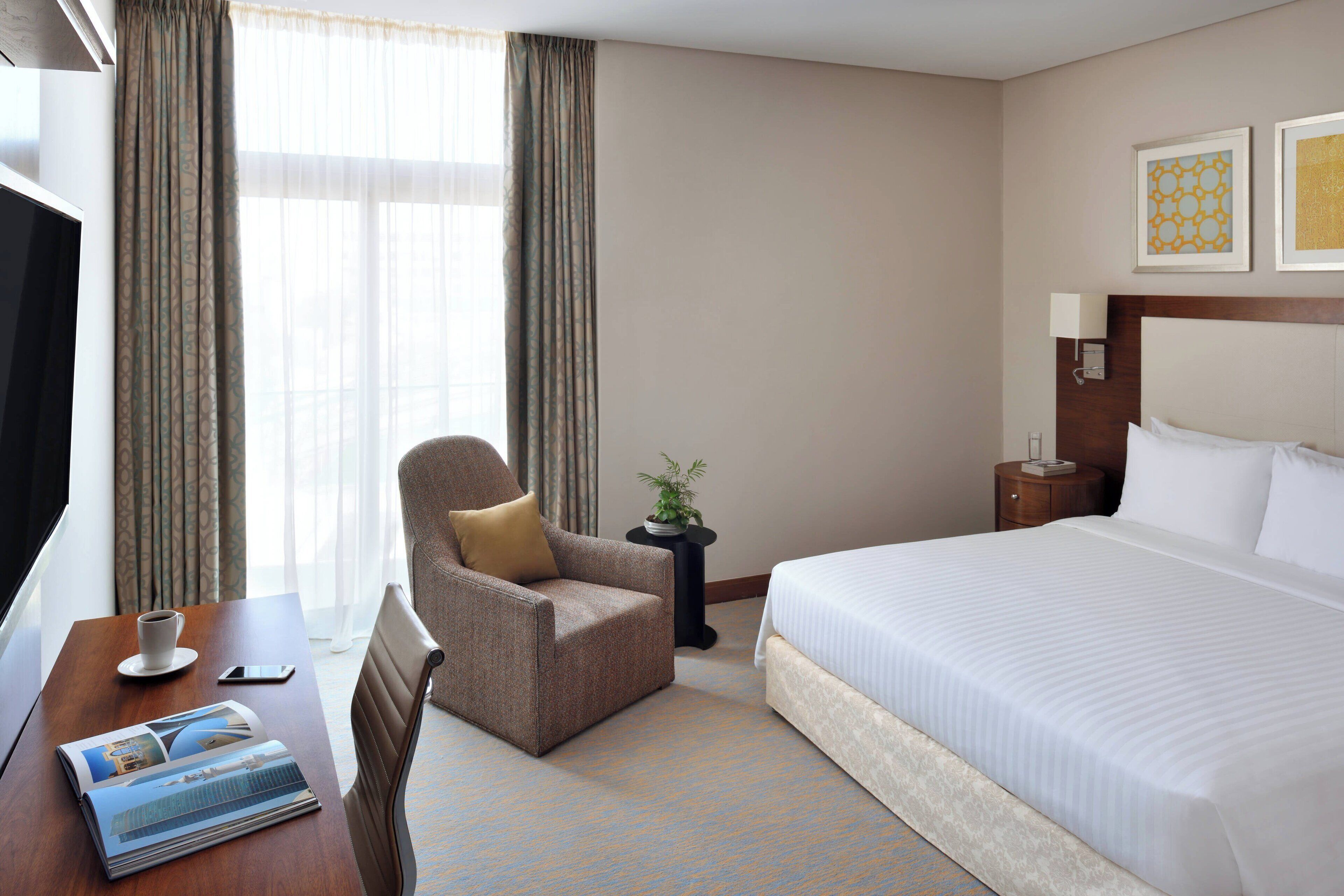 Quarto de hotel com cama, cadeira, escrivaninha com telefone e revista, televisão e janela com cortinas.