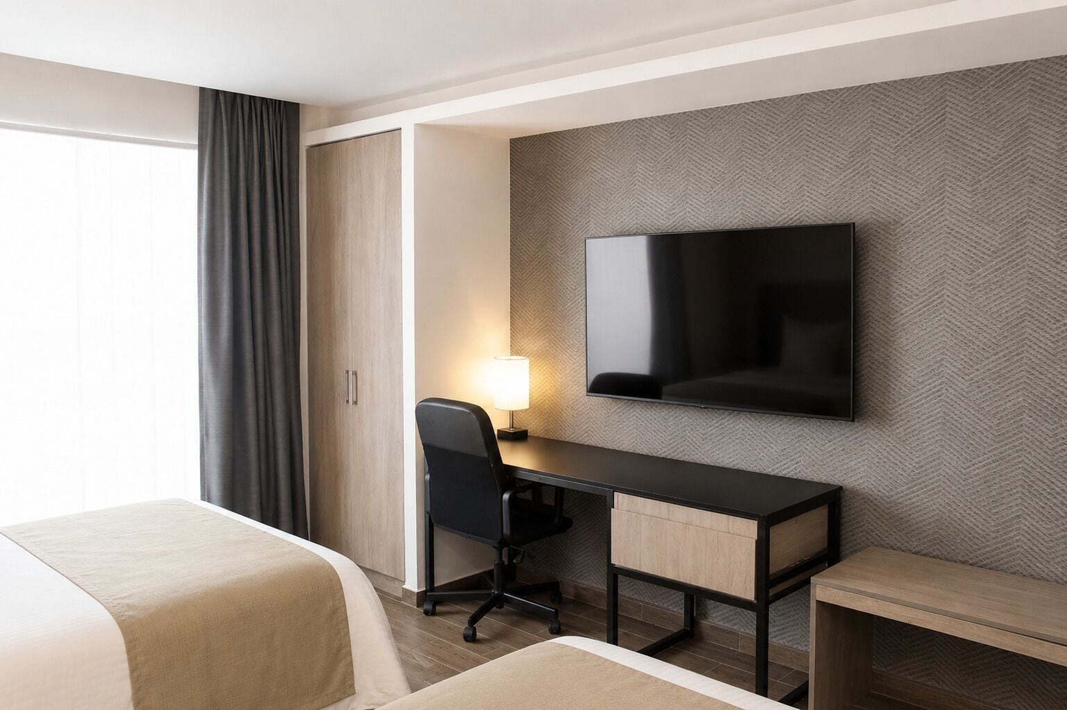 Quarto de hotel com cama, escrivaninha com cadeira, televisão e guarda-roupa.