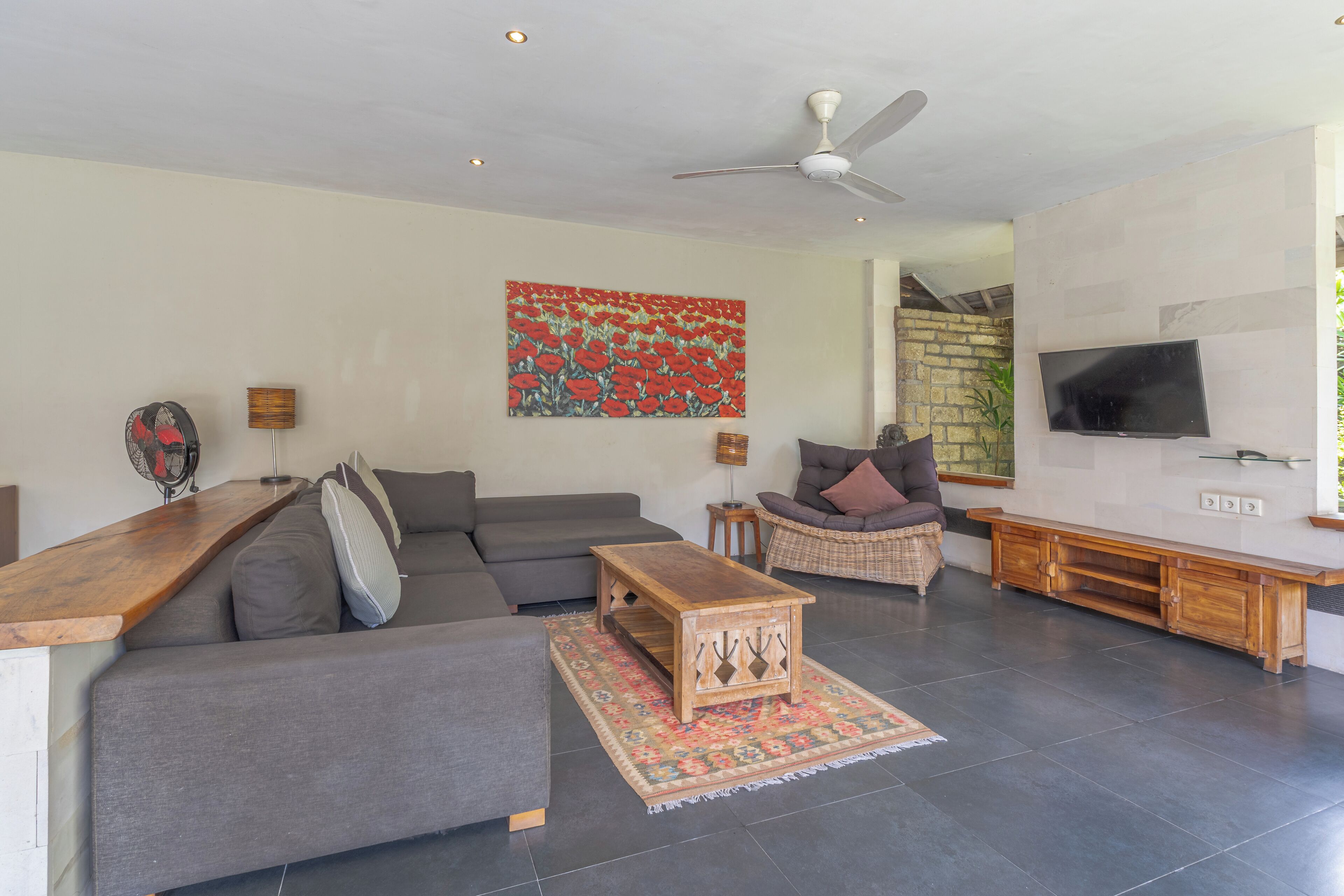 Uma sala de estar moderna com um sofá cinza, uma mesa de centro de madeira, uma TV fixada na parede e um ventilador de teto.