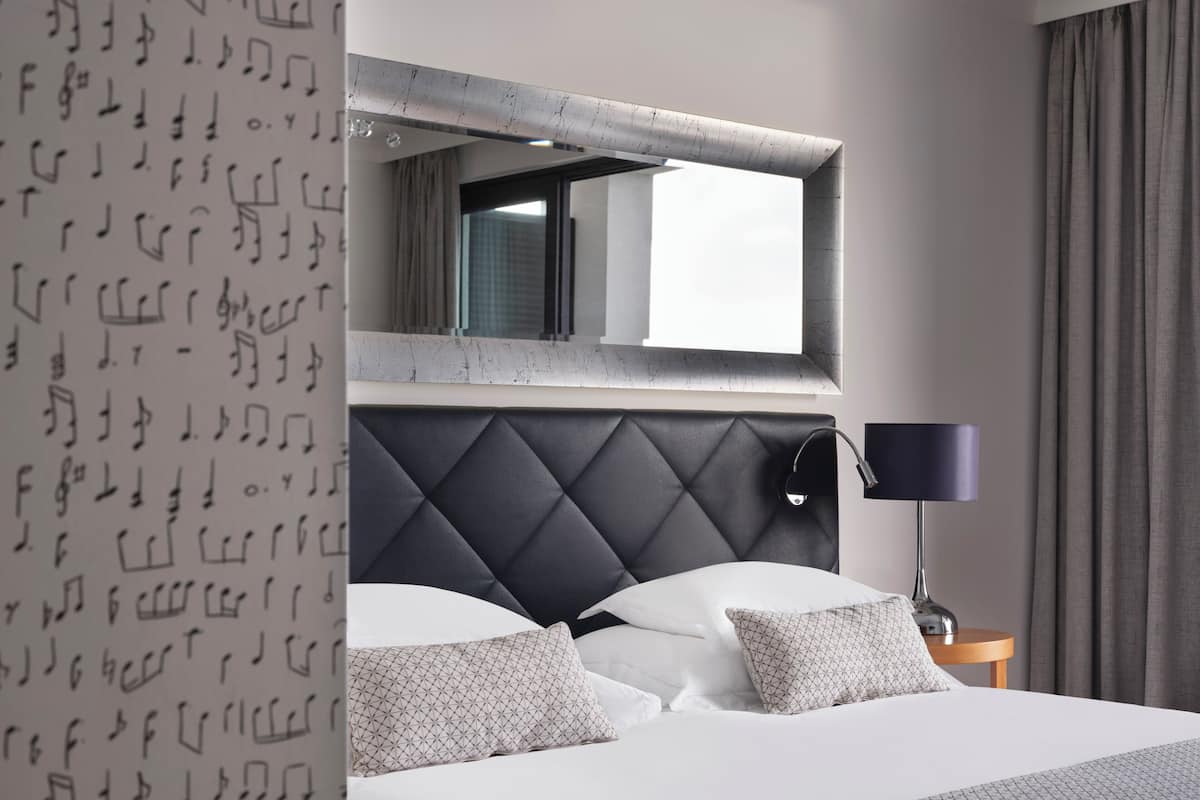 Quarto de hotel com cama, mesa de cabeceira, luminária, espelho e janela com cortinas.
