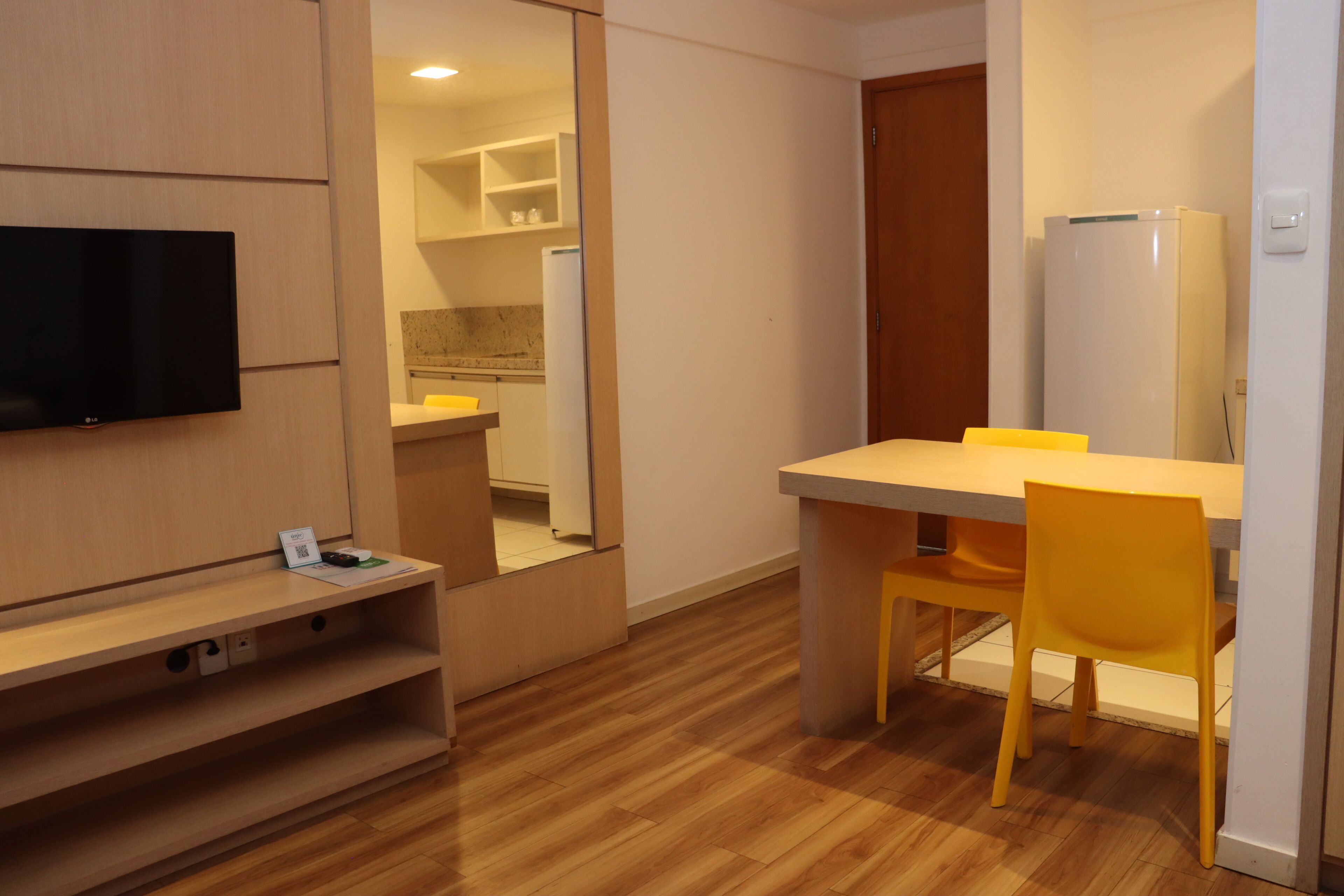 Cozinha moderna com piso de madeira, televisão embutida e área de jantar com mesa e cadeiras amarelas.
