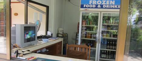Uma pequena loja fechada com um letreiro indicando 'FROZEN FOOD & DRINKS'. No interior, há uma geladeira repleta de bebidas, uma televisão sobre uma mesa e uma cadeira.