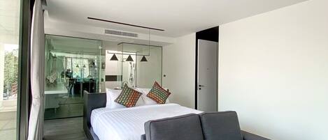 Quarto de hotel moderno com uma cama grande, um sofá cinzento e um separador de vidro dividindo o quarto de dormir do banheiro.