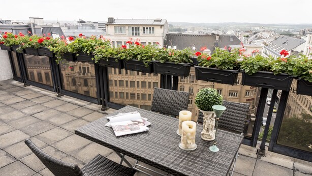 Terraço no telhado com uma mesa posta para dois, rodeado por cadeiras de vime e decorado com flores vermelhas em vasos pretos.