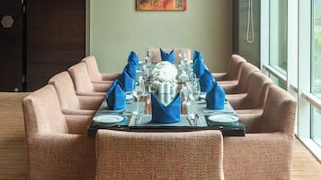 Uma sala de jantar com uma mesa posta para quatro pessoas, rodeada por cadeiras estofadas, uma janela grande e um teto com iluminação embutida.