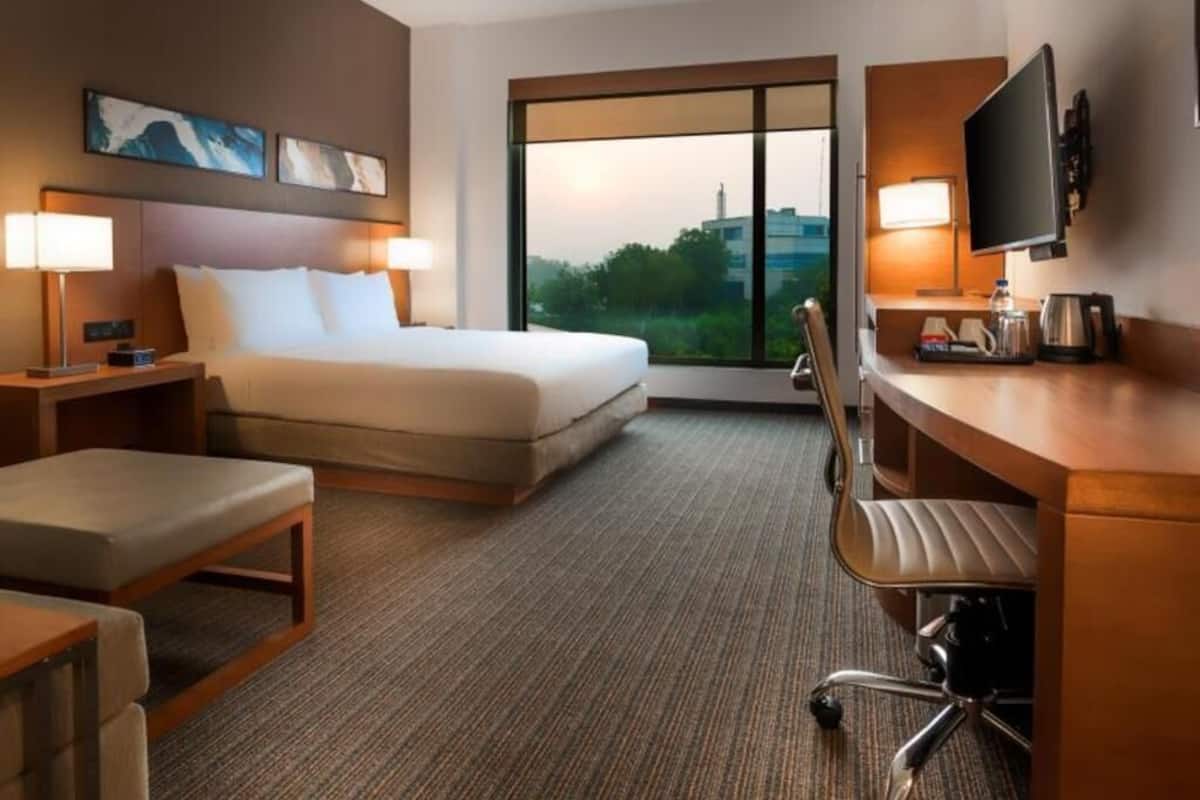 Quarto de hotel com cama, escrivaninha, cadeira, luminária, televisão e uma grande janela com vista.