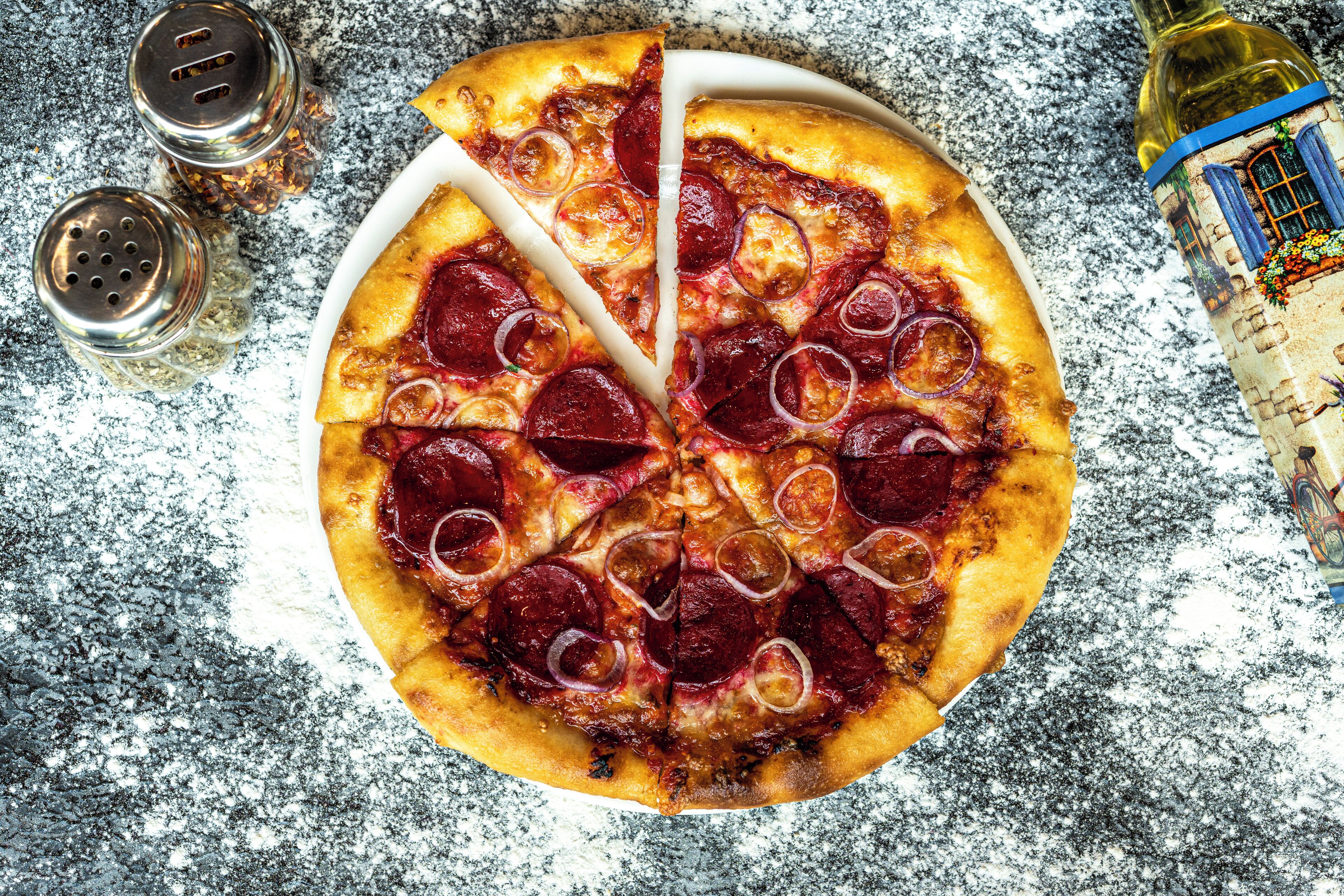 Uma pizza de pepperoni em um prato branco com uma fatia retirada, cercada por saleiros e pimenteiros, e uma garrafa de azeite sobre uma bancada de granito.