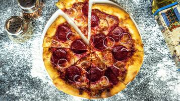 Uma pizza de pepperoni em um prato branco com uma fatia retirada, cercada por saleiros e pimenteiros, e uma garrafa de azeite sobre uma bancada de granito.