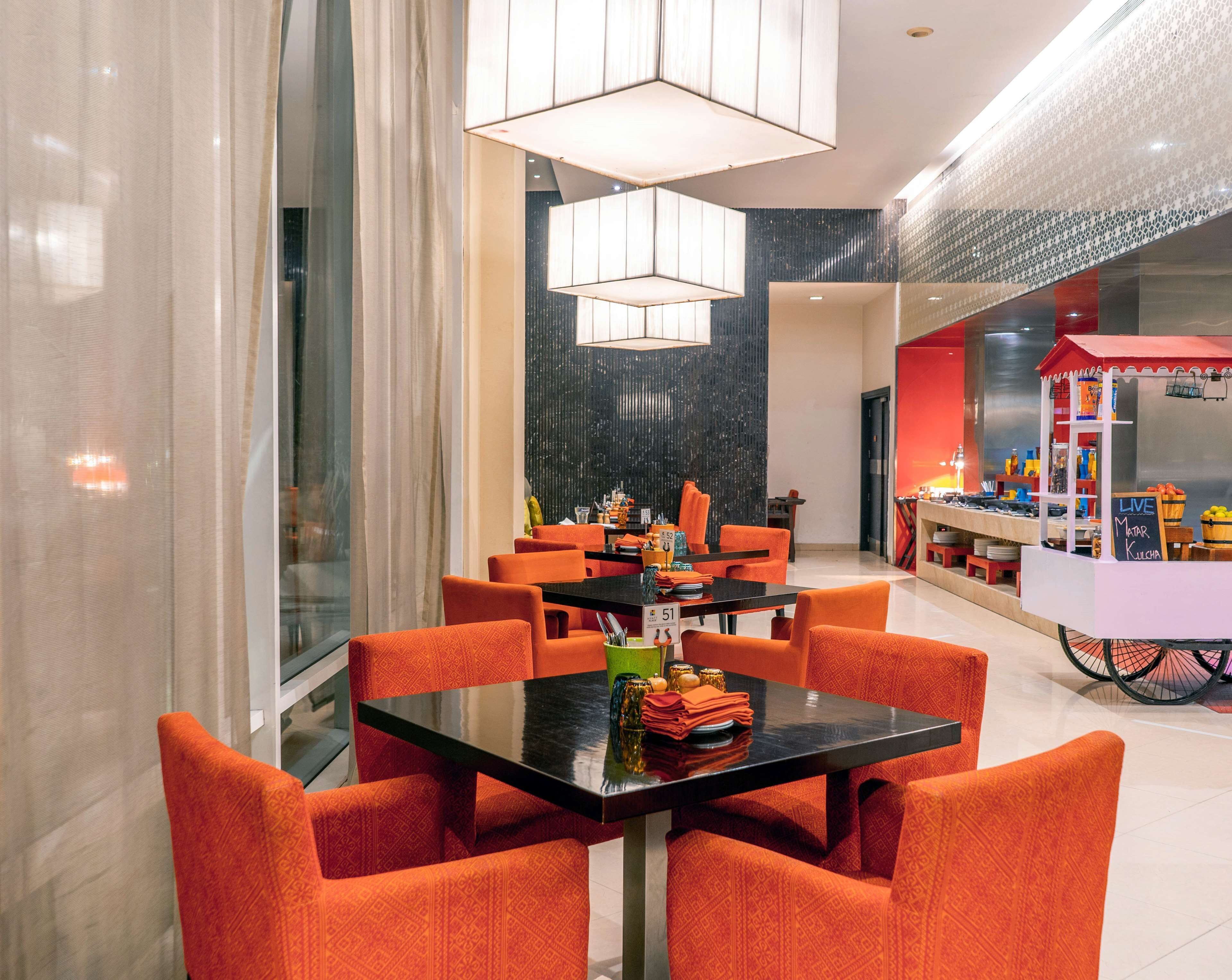 Uma área de refeições moderna com cadeiras laranja e uma mesa preta, destacando um lustre central e um quiosque de comida ao fundo.