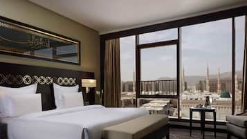 Quarto de hotel com uma cama grande, um sofá pequeno, uma mesa e vista para um horizonte urbano com uma mesquita.