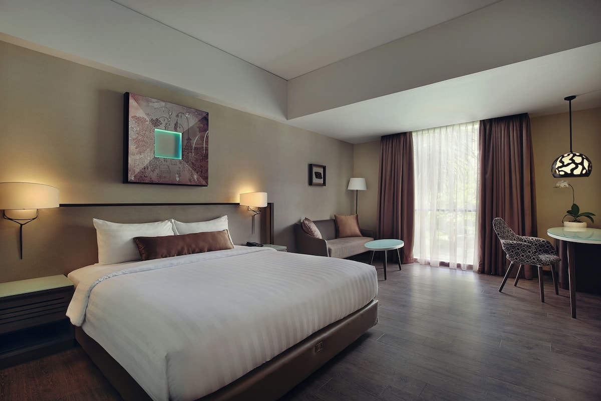 Quarto de hotel com cama grande, escrivaninha, cadeira e mesa pequena.