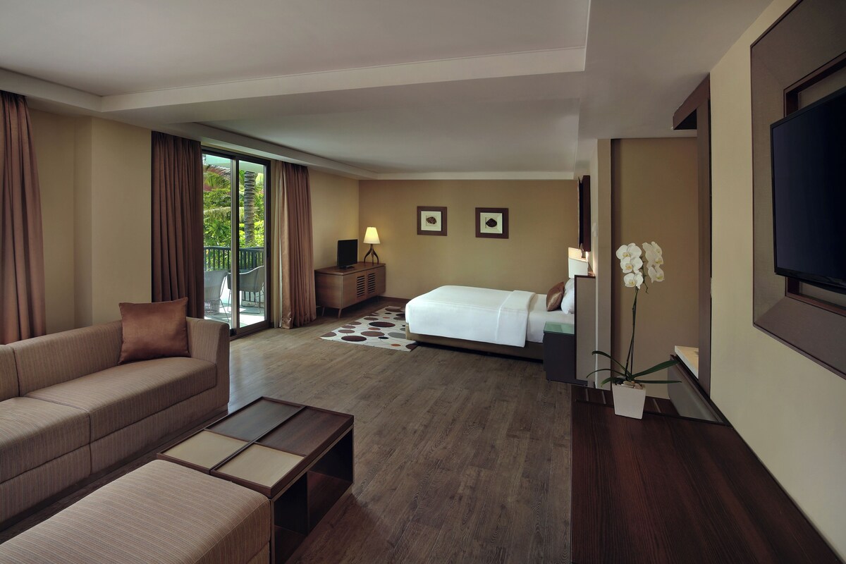 Quarto de hotel com sofá, cama, televisão e varanda com cortinas.