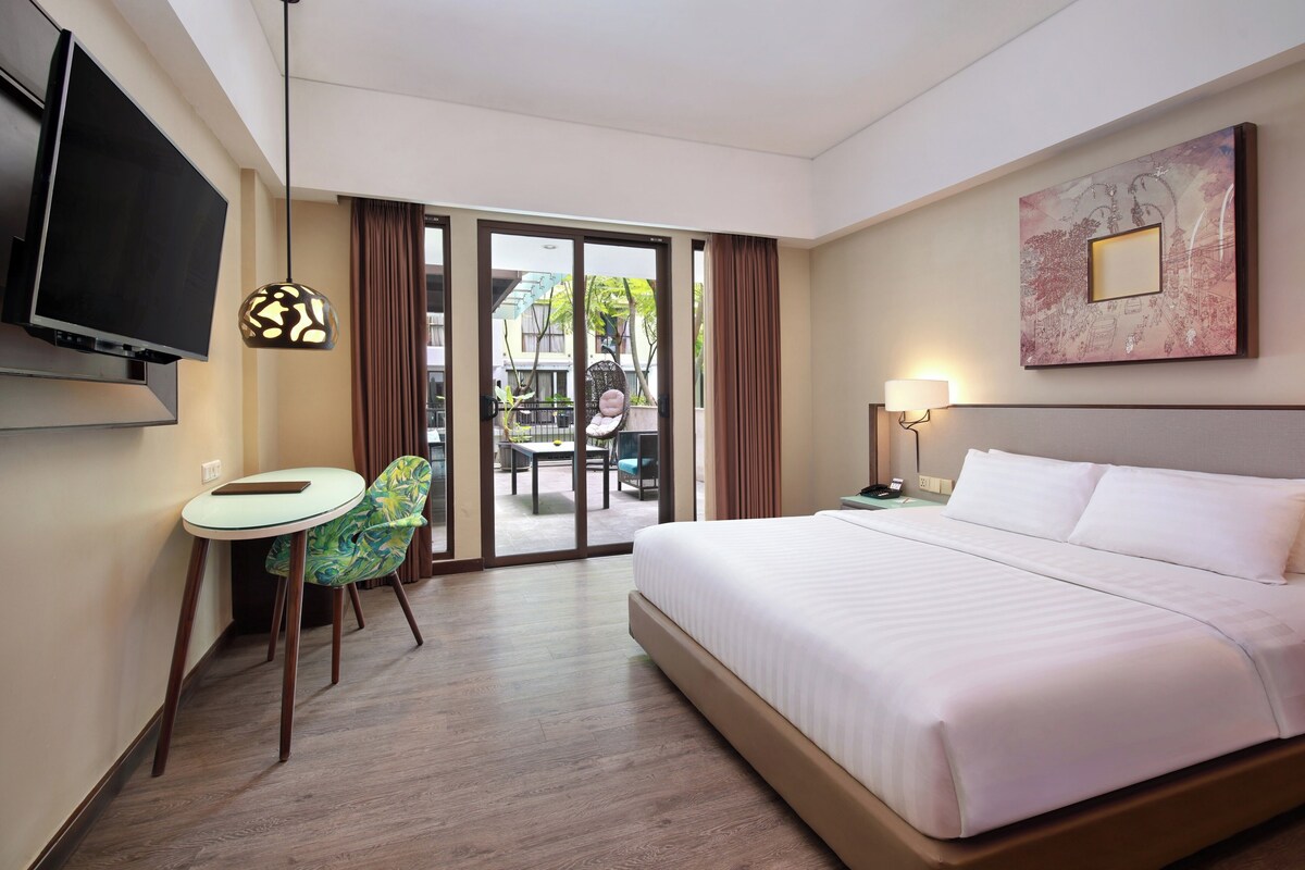 Quarto de hotel com cama, televisão, mesa com cadeira e vista para o exterior através de uma porta deslizante.