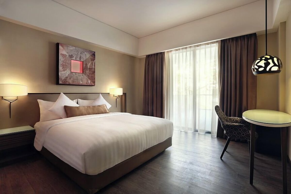 Quarto de hotel com uma cama grande, duas luminárias de cabeceira, uma cadeira, uma mesinha e uma janela ampla com cortinas.