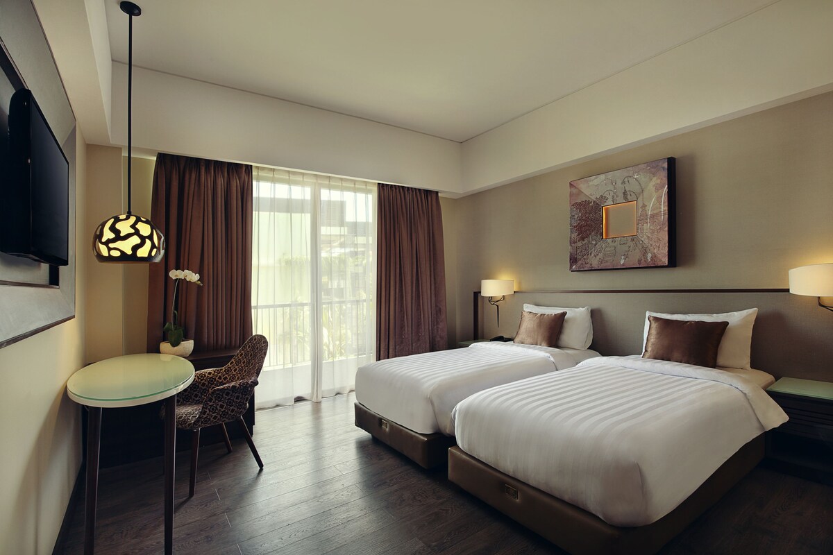 Quarto de hotel com duas camas, uma escrivaninha, uma cadeira, televisão e janela com cortinas.