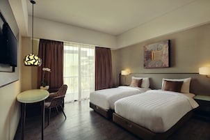 Quarto de hotel com duas camas, uma escrivaninha, uma cadeira, televisão e janela com cortinas.